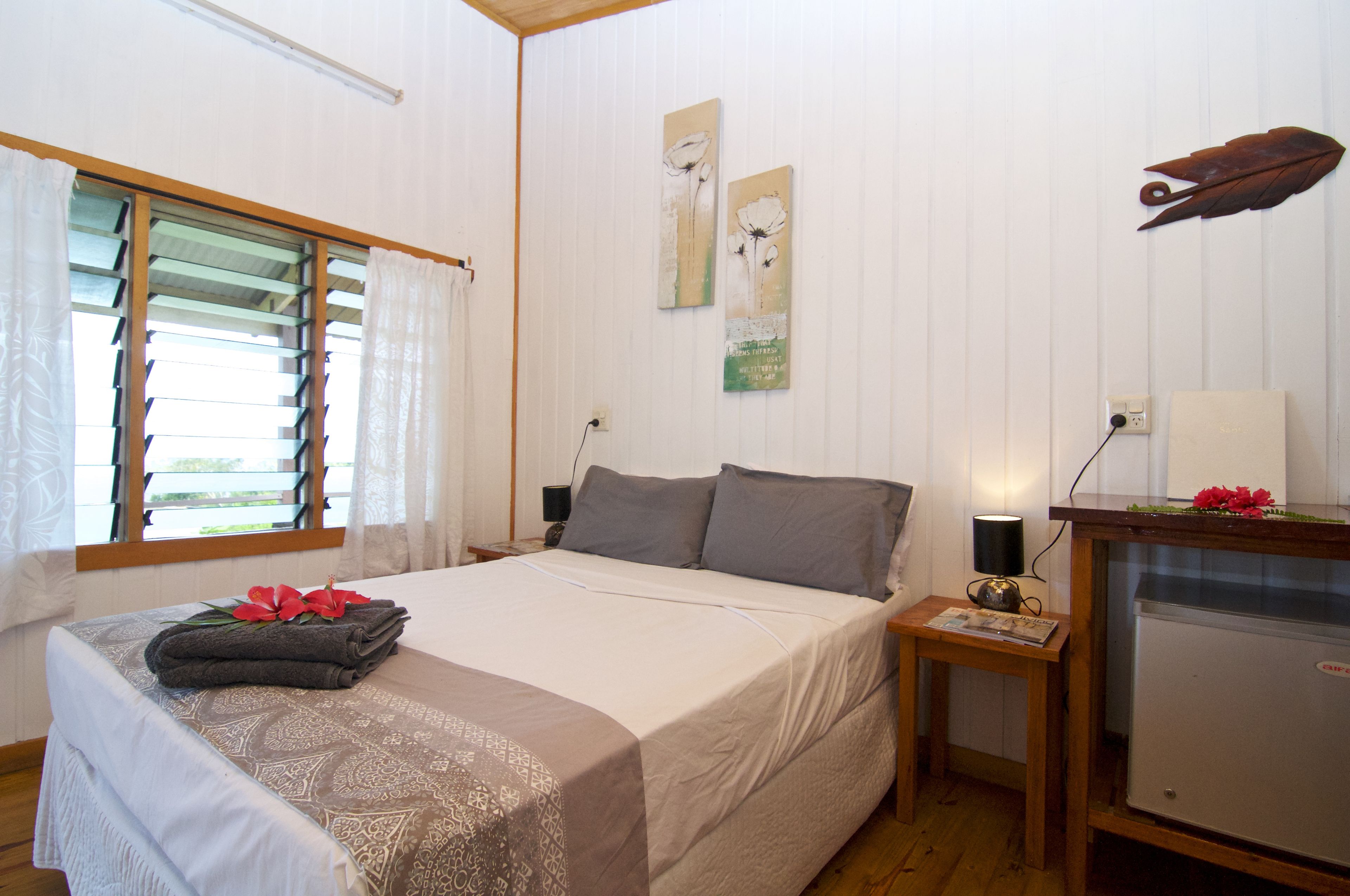 Uma cama bem arrumada com um travesseiro decorativo e uma toalha dobrada, uma mesinha de cabeceira com abajur, um pequeno frigorífico e uma janela com persianas.