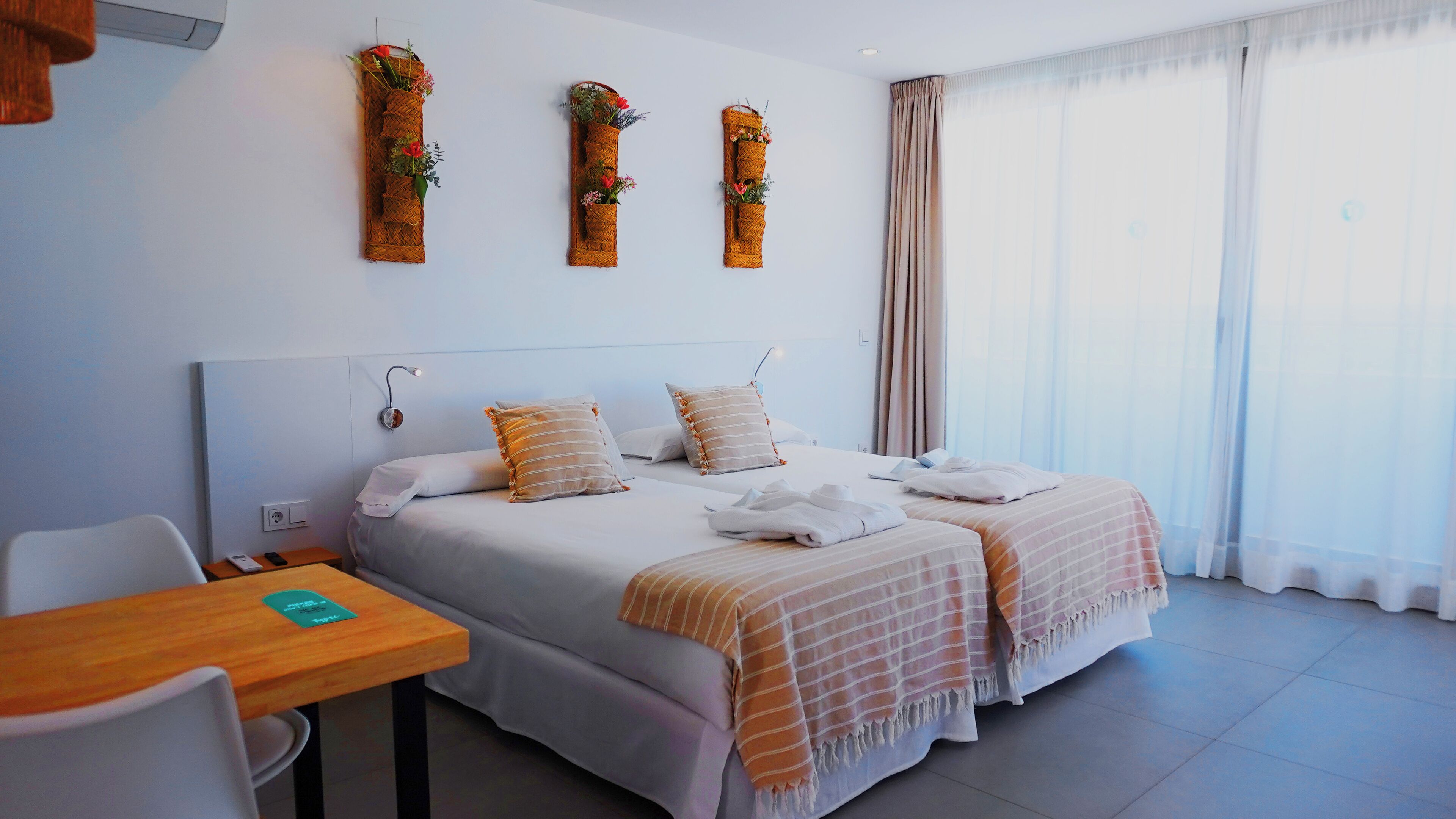 Quarto de hotel com uma cama, duas almofadas, uma mesa de cabeceira, uma cadeira e um candeeiro fixado na parede.