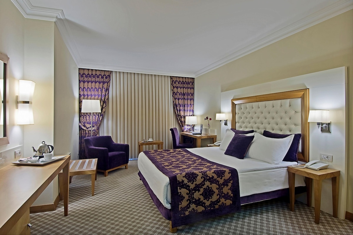 Quarto de hotel com uma cama grande, duas cadeiras, uma escrivaninha e uma mesa de cabeceira.