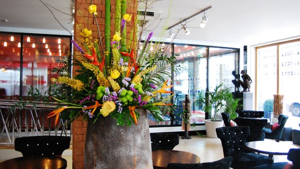 Um espaço interno moderno com um arranjo floral grande, cadeiras estofadas em preto e uma grande janela com vista para o exterior.