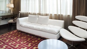 Quarto de hotel moderno com sofá branco, tapete estampado, mesa redonda e janela grande com cortinas translúcidas.