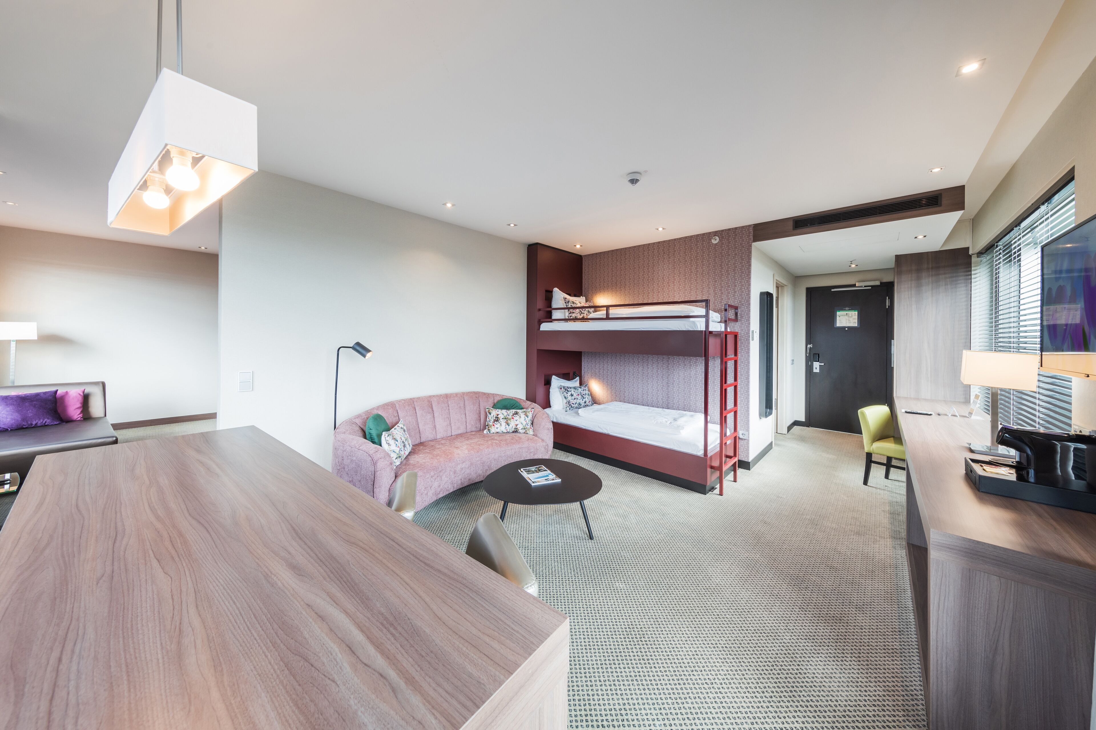 Quarto de hotel moderno com beliche, sofá, mesa de jantar e escrivaninha.
