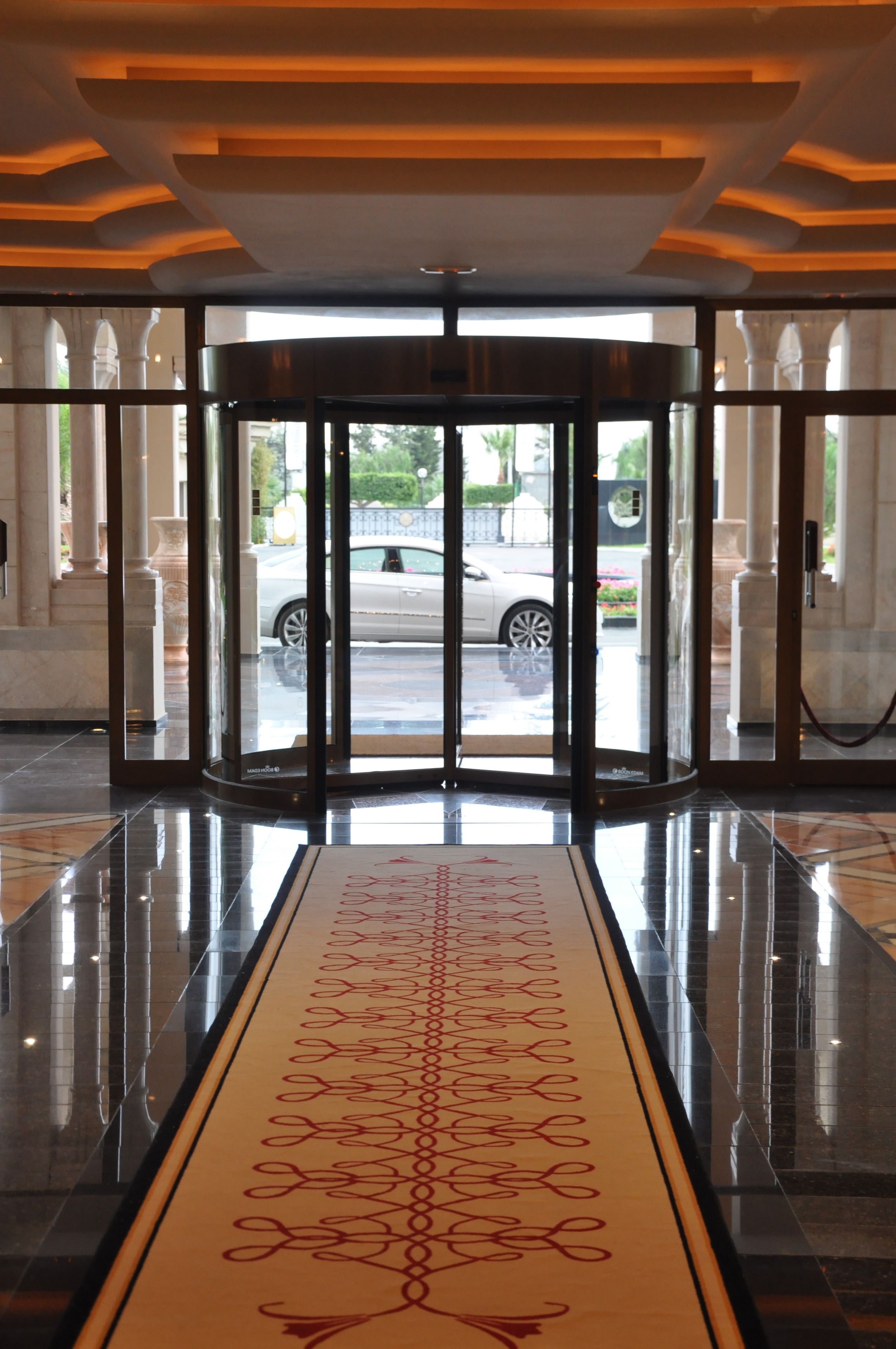 Um hall de hotel com piso de mármore, uma coluna central e vista para um carro estacionado lá fora.