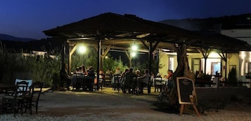 Uma área de refeições ao ar livre com um pavilhão coberto, mesas, cadeiras e algumas pessoas sentadas.