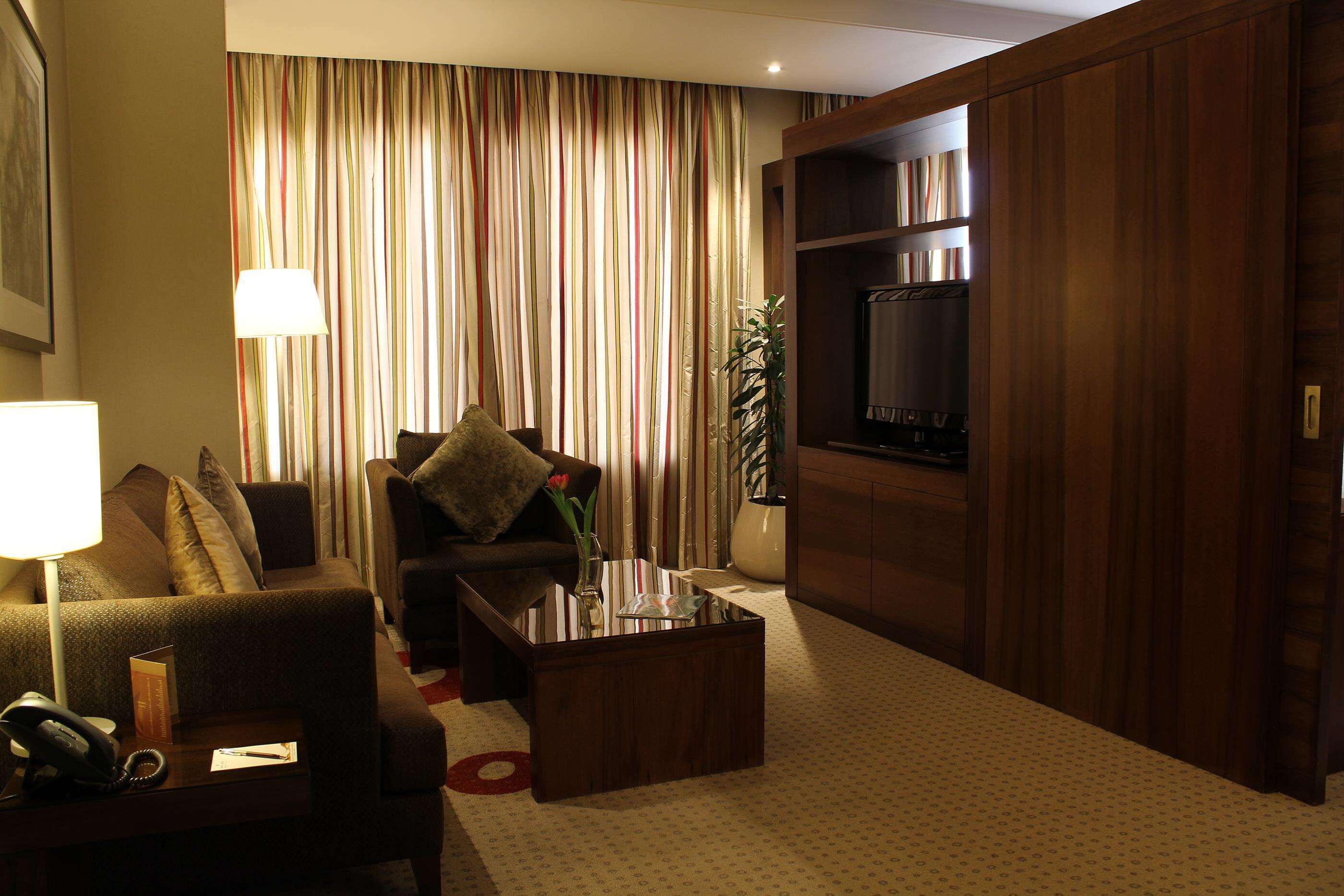 Quarto de hotel com sofá, mesa de centro, luminária, televisão e cortina.