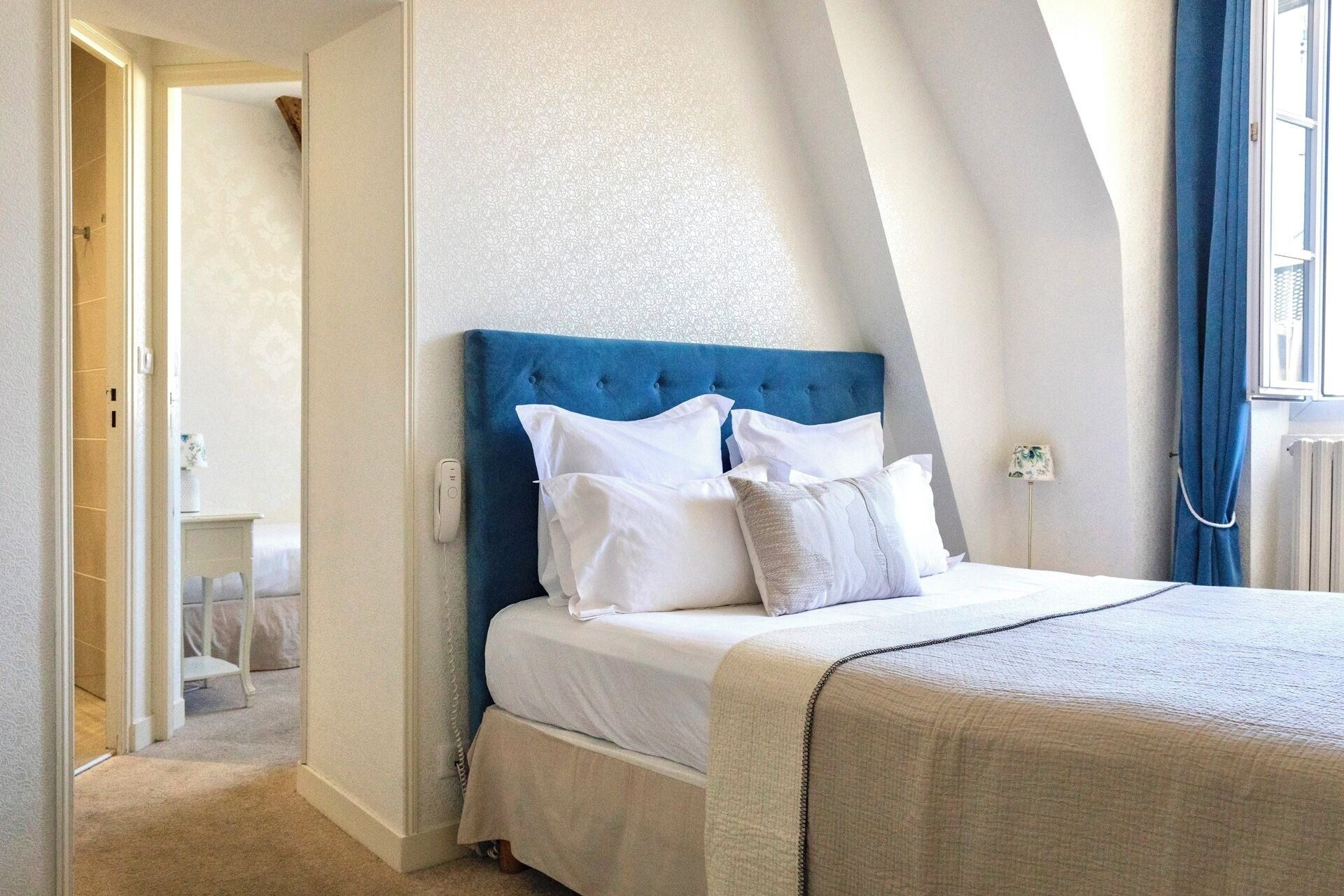 Quarto com uma cama grande, cabeceira azul, roupa de cama branca e vista para o exterior pela janela.