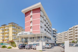 Um hotel com vários andares, fachada marcante em vermelho e branco, entrada coberta e área de estacionamento com vários carros.