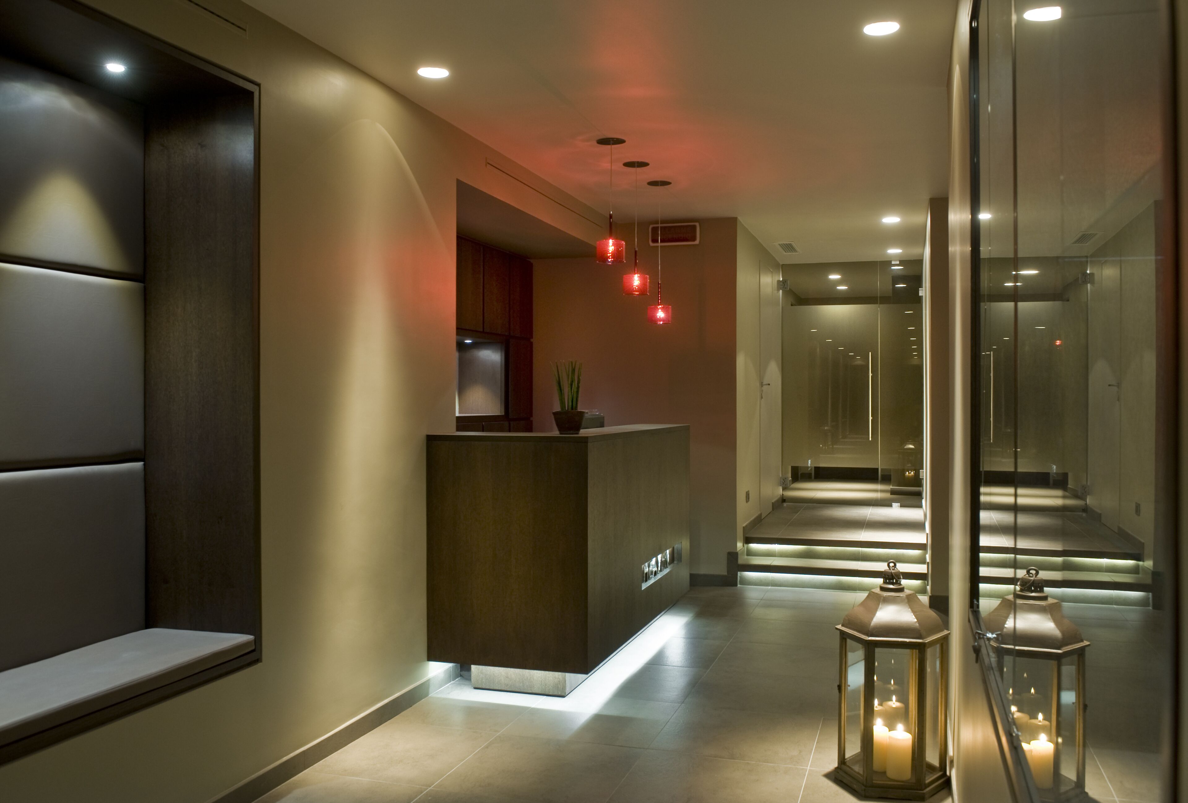 Uma área de recepção de hotel moderna, com um balcão de madeira, uma planta em vaso e luminárias pendentes vermelhas.