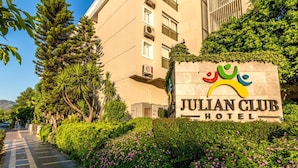 Hotel Julian Club, com uma placa bem visível, cercado por vegetação exuberante e céu claro.