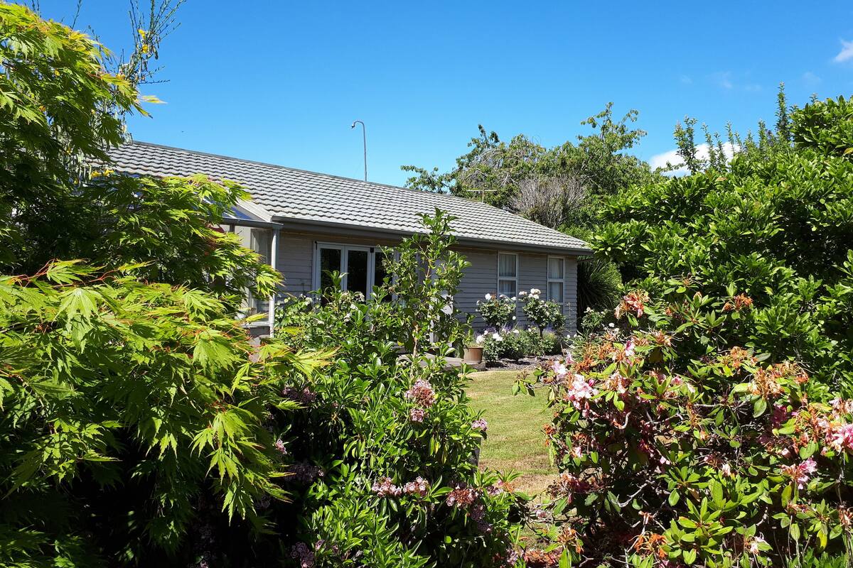Uma casa com telhado de telhas, rodeada por vegetação exuberante e plantas floridas.