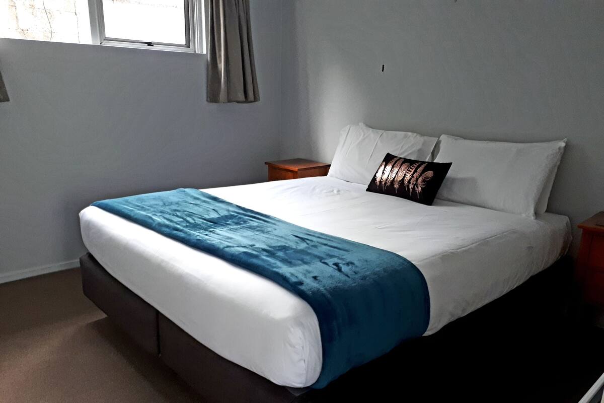 Uma cama bem arrumada com colcha branca e azul, um travesseiro preto com desenho de pena e uma mesinha de cabeceira de madeira ao lado.