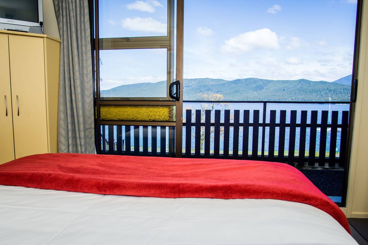 Quarto com cama, janela com vista para montanhas e um lago, e um guarda-roupa amarelo.