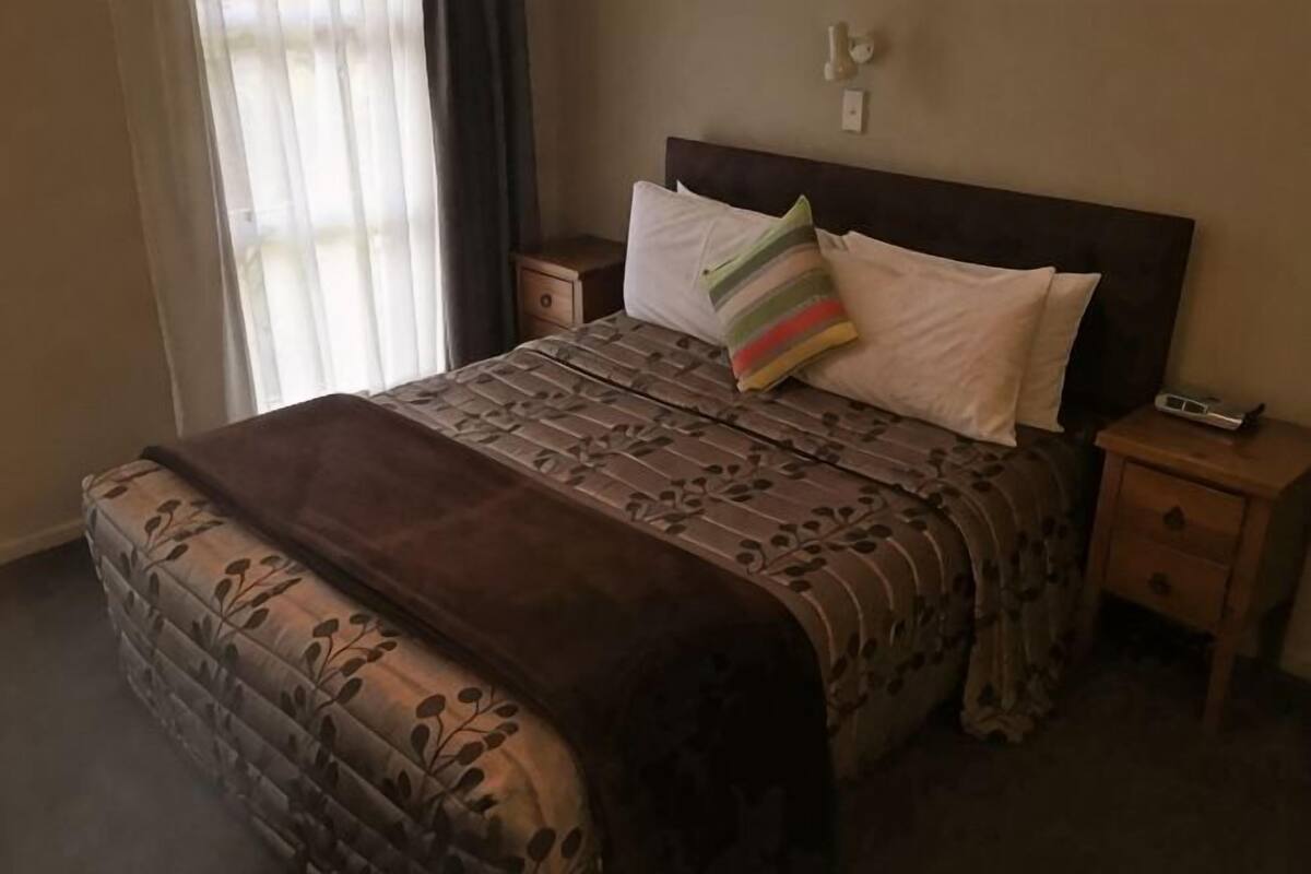 Uma cama bem arrumada com colcha estampada, uma mesa de cabeceira com abajur e uma janela com cortinas.