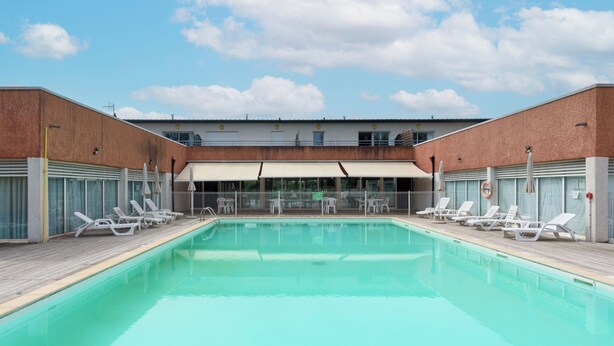 Piscina com espreguiçadeiras e guarda-sóis, rodeada por um edifício com telhado plano e janelas amplas.