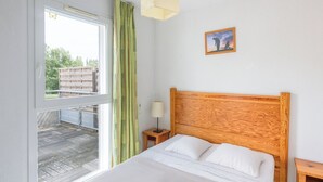 Quarto com cabeceira de madeira, cama com roupa de cama branca, mesa de cabeceira com luminária e janela com vista para um deck de madeira e vegetação.