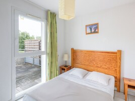 Quarto com cabeceira de madeira, cama com roupa de cama branca, mesa de cabeceira com luminária e janela com vista para um deck de madeira e vegetação.