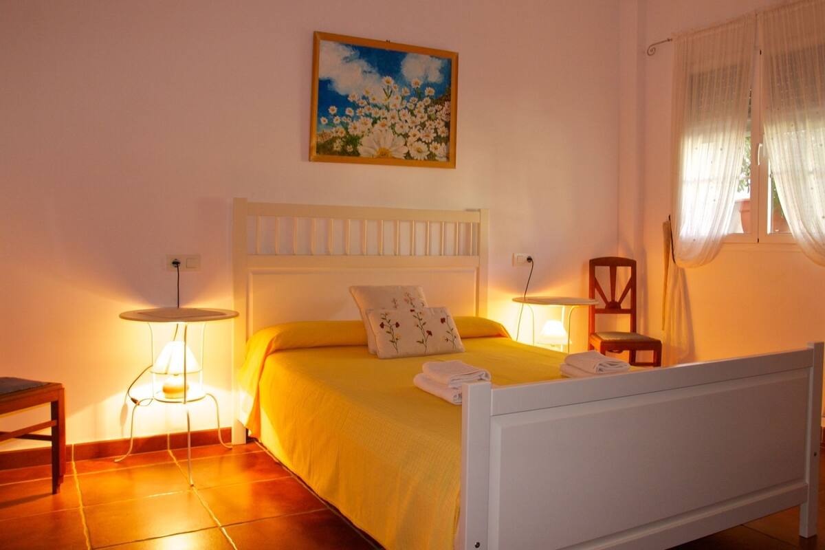 Um quarto com cama, mesas de cabeceira, uma cadeira, um quadro e uma janela com cortinas.