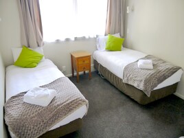 Quarto de hotel com duas camas de solteiro, cada uma com travesseiro e cobertor. Há uma pequena mesa de cabeceira de madeira entre as camas.