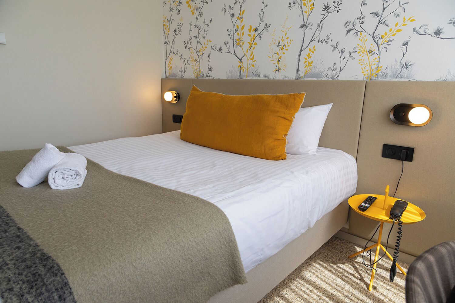 Quarto de hotel com uma cama, mesa de cabeceira com telefone e parede com papel de parede floral.