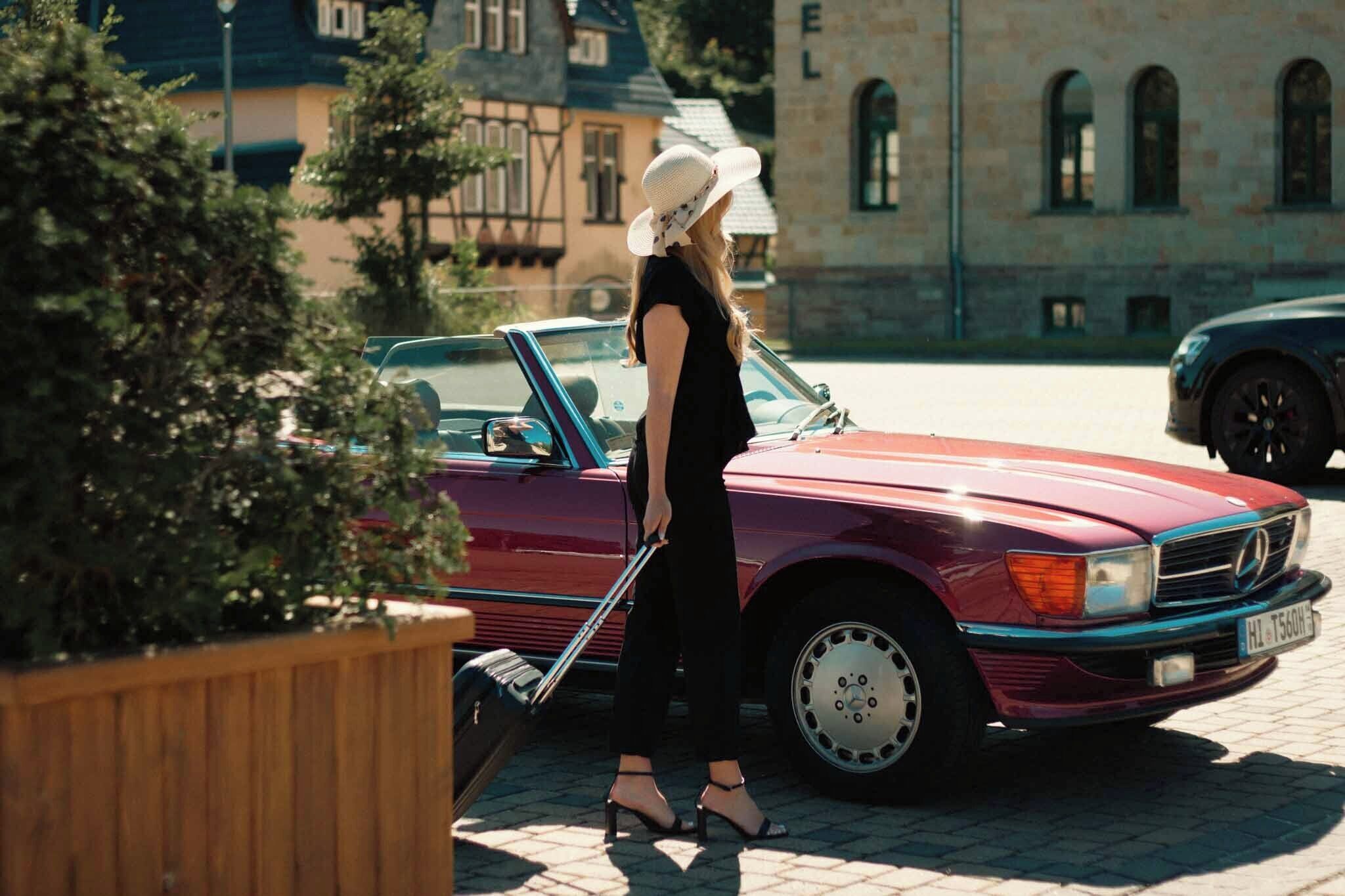 Uma mulher de vestido preto e chapéu branco caminhando perto de um carro conversível vermelho estacionado em uma rua de paralelepípedos.