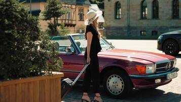 Uma mulher de vestido preto e chapéu branco caminhando perto de um carro conversível vermelho estacionado em uma rua de paralelepípedos.