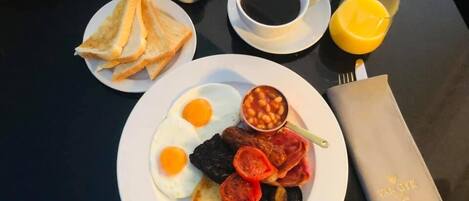Prato de café da manhã com ovos, bacon, tomates, feijão e torradas em uma mesa com uma xícara de café, suco e um guardanapo.