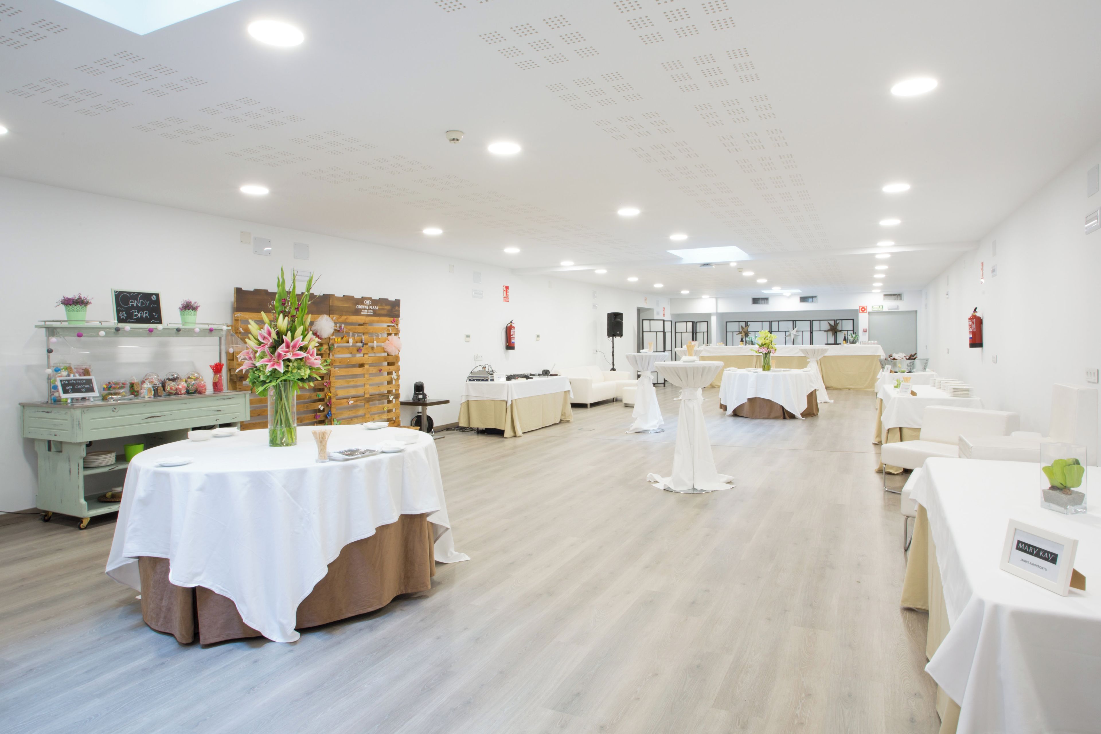 Um salão de eventos espaçoso com várias mesas redondas cobertas por toalhas brancas, uma área de palco e disposição para assentos.