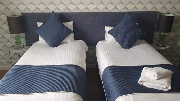 Quarto de hotel com duas camas, cada uma com cabeceira azul, roupa de cama branca e almofada de destaque azul. Há duas luminárias de cabeceira e uma pequena mesa com uma planta verde.