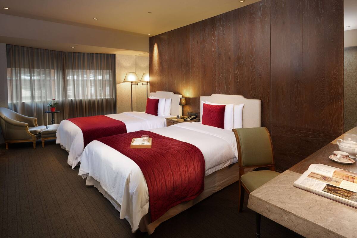 Quarto de hotel com duas camas, uma escrivaninha, uma cadeira e uma luminária.