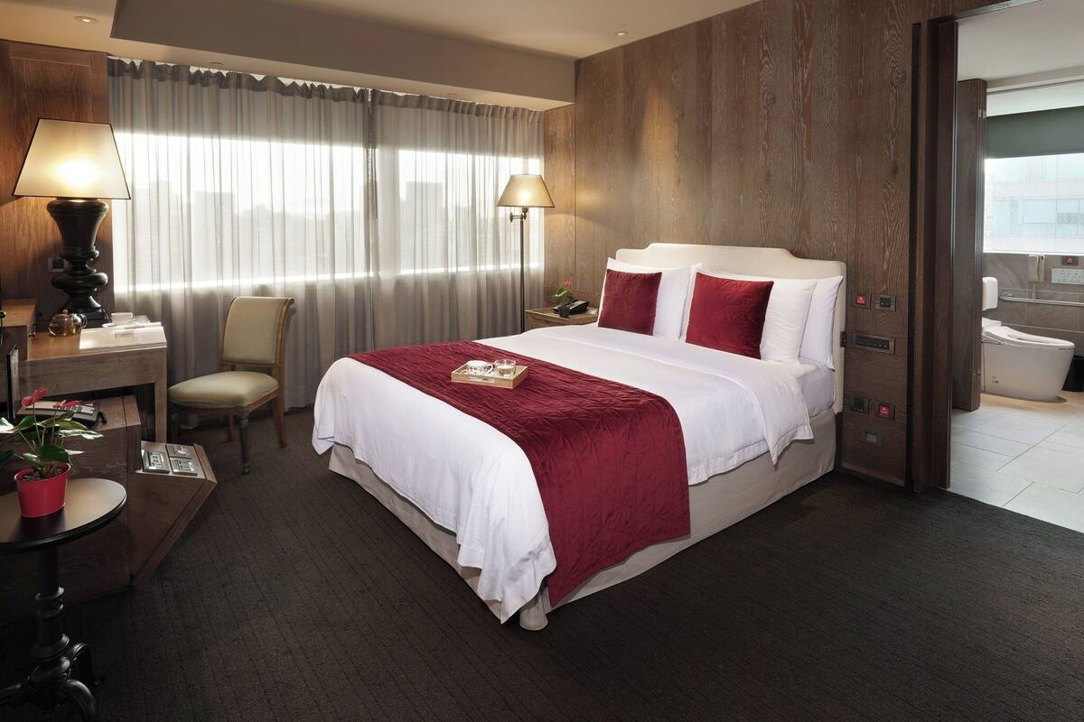 Quarto de hotel com uma cama grande, duas luminárias de cabeceira, uma escrivaninha com cadeira, uma mesinha com planta em vaso, e vista para o banheiro.