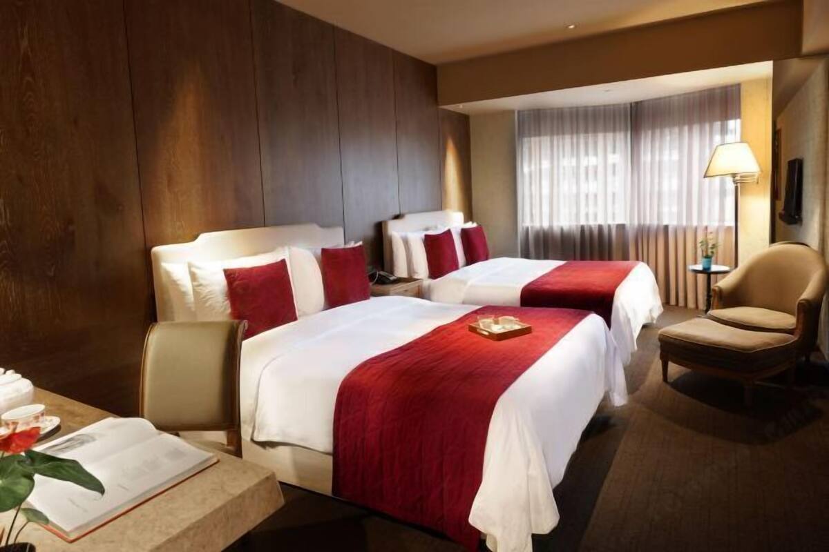 Quarto de hotel com duas camas, uma escrivaninha com cadeira, uma luminária e vista para o exterior.