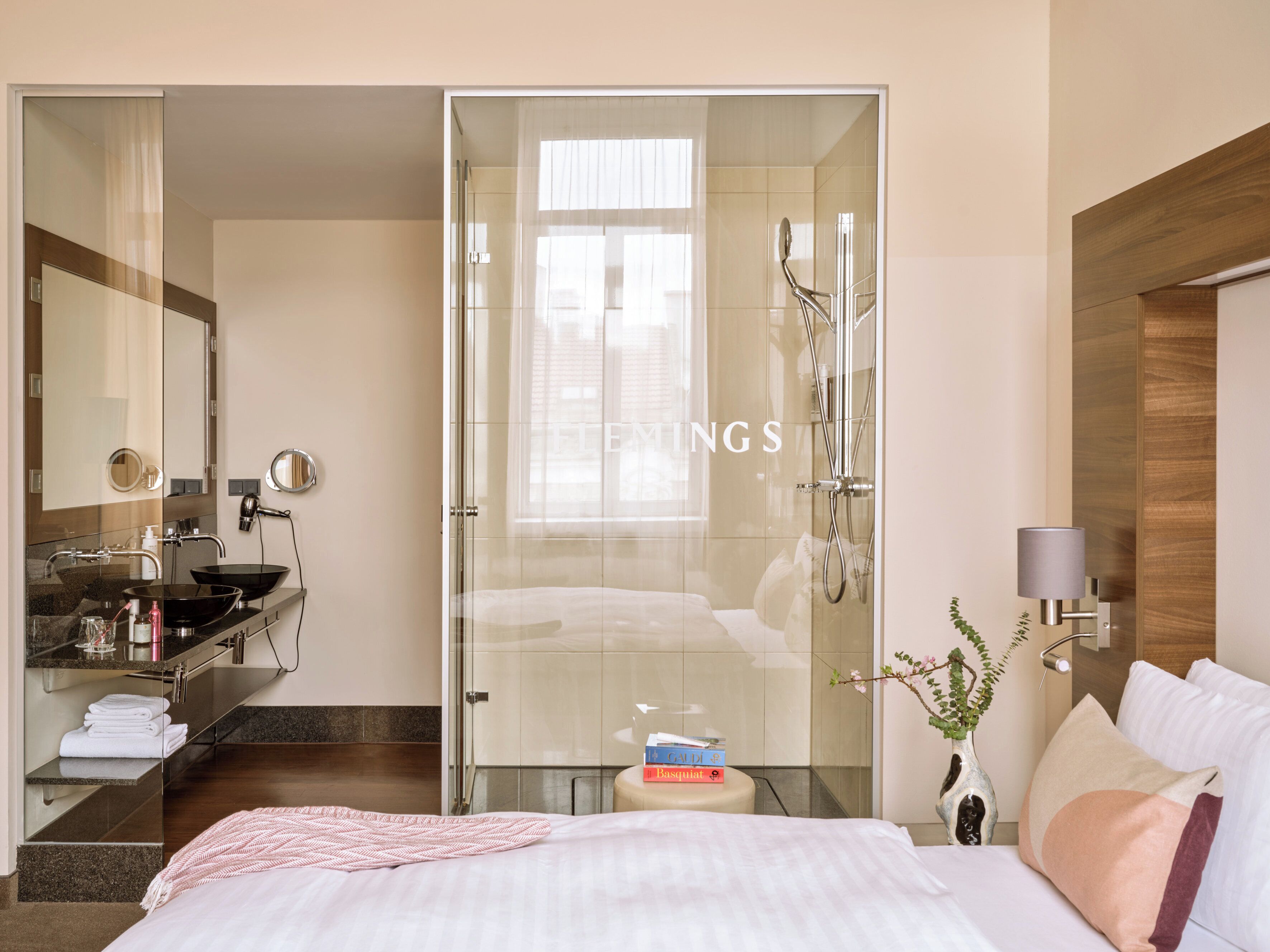 Quarto de hotel moderno com chuveiro em gabinete de vidro, toucador com cuba preta, cama com roupa de cama branca e mesa de cabeceira com luminária.