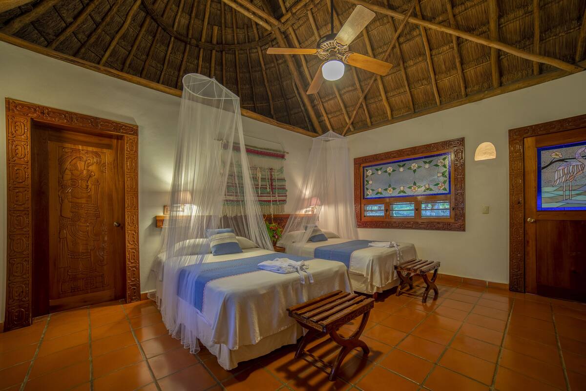 Quarto com duas camas, um banco de madeira, um ventilador de teto e uma porta decorativa.