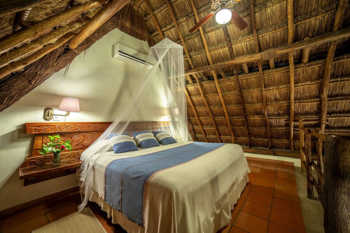 Quarto com cama sob telhado de palha, mesa de cabeceira de madeira com abajur e uma pequena planta.