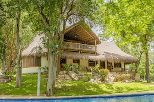Uma casa com telhado de palha e varanda, rodeada por vegetação exuberante e uma piscina.