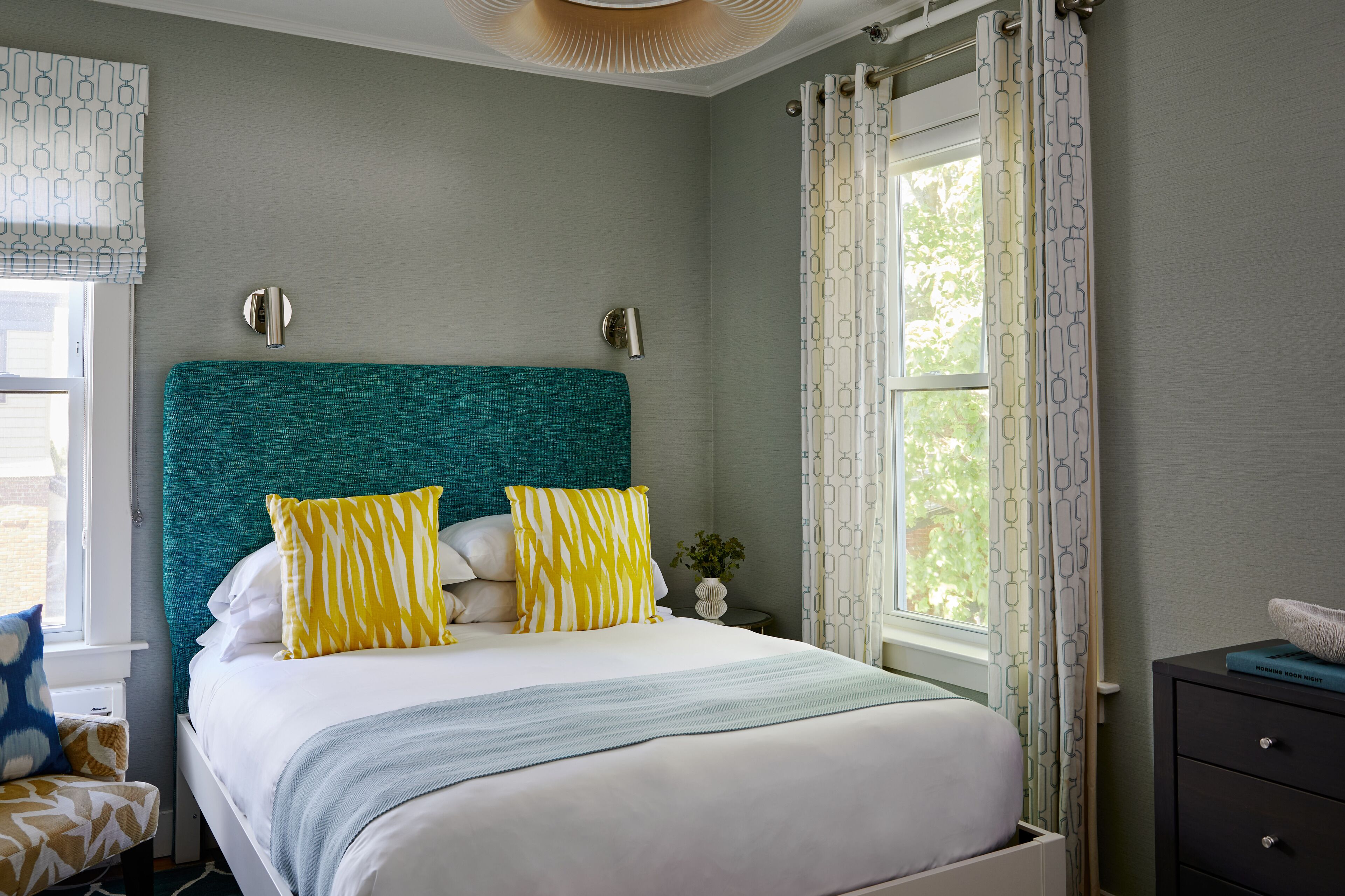 Quarto com cama, mesa de cabeceira, janela com cortinas e vista para o verde lá fora.