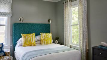 Quarto com cama, mesa de cabeceira, janela com cortinas e vista para o verde lá fora.