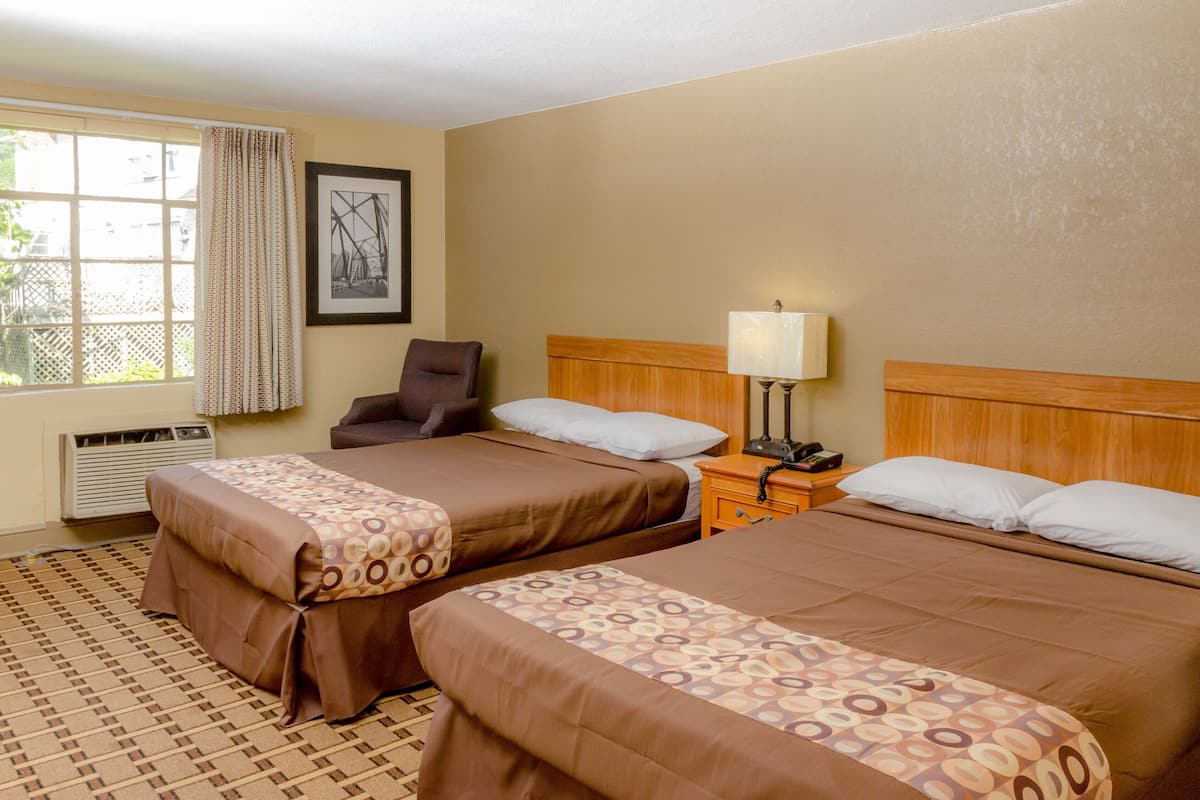 Quarto de hotel com duas camas, uma cadeira, mesa de cabeceira com abajur e janela com cortinas.