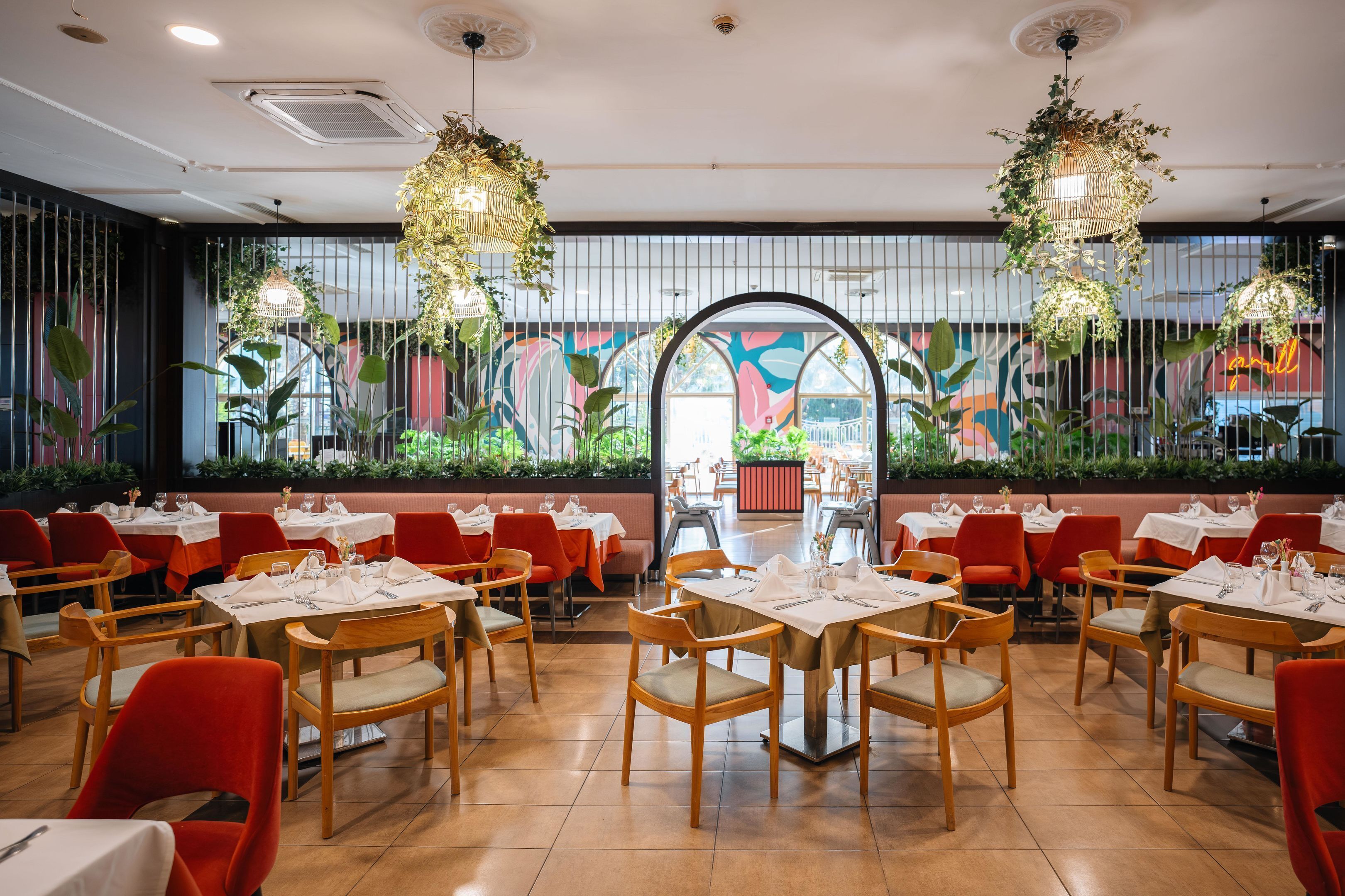 Um restaurante com várias mesas e cadeiras, um mural colorido na parede e plantas penduradas.