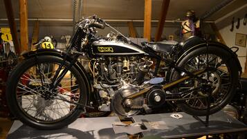 Uma motocicleta antiga em exposição em uma garagem com vigas de madeira e diversas ferramentas e equipamentos ao fundo.