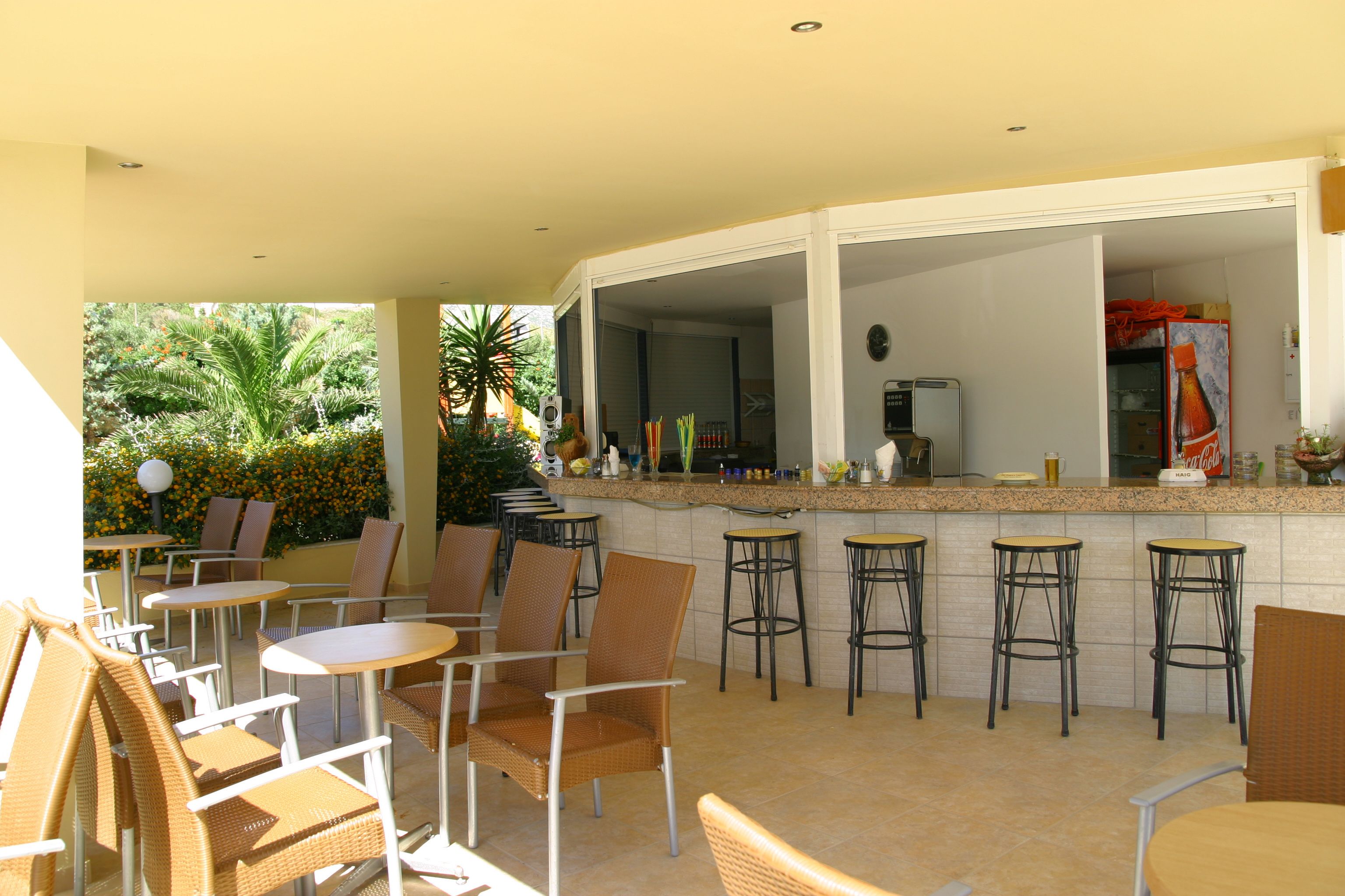 Uma área de pátio ao ar livre com cadeiras e mesas de vime, um bar com banquetas e vista para o jardim.
