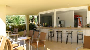 Uma área de pátio ao ar livre com cadeiras e mesas de vime, um bar com banquetas e vista para o jardim.