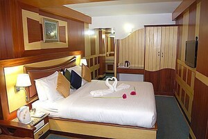 Quarto de hotel com uma cama bem arrumada, mesa de cabeceira com abajur, televisão e guarda-roupa.