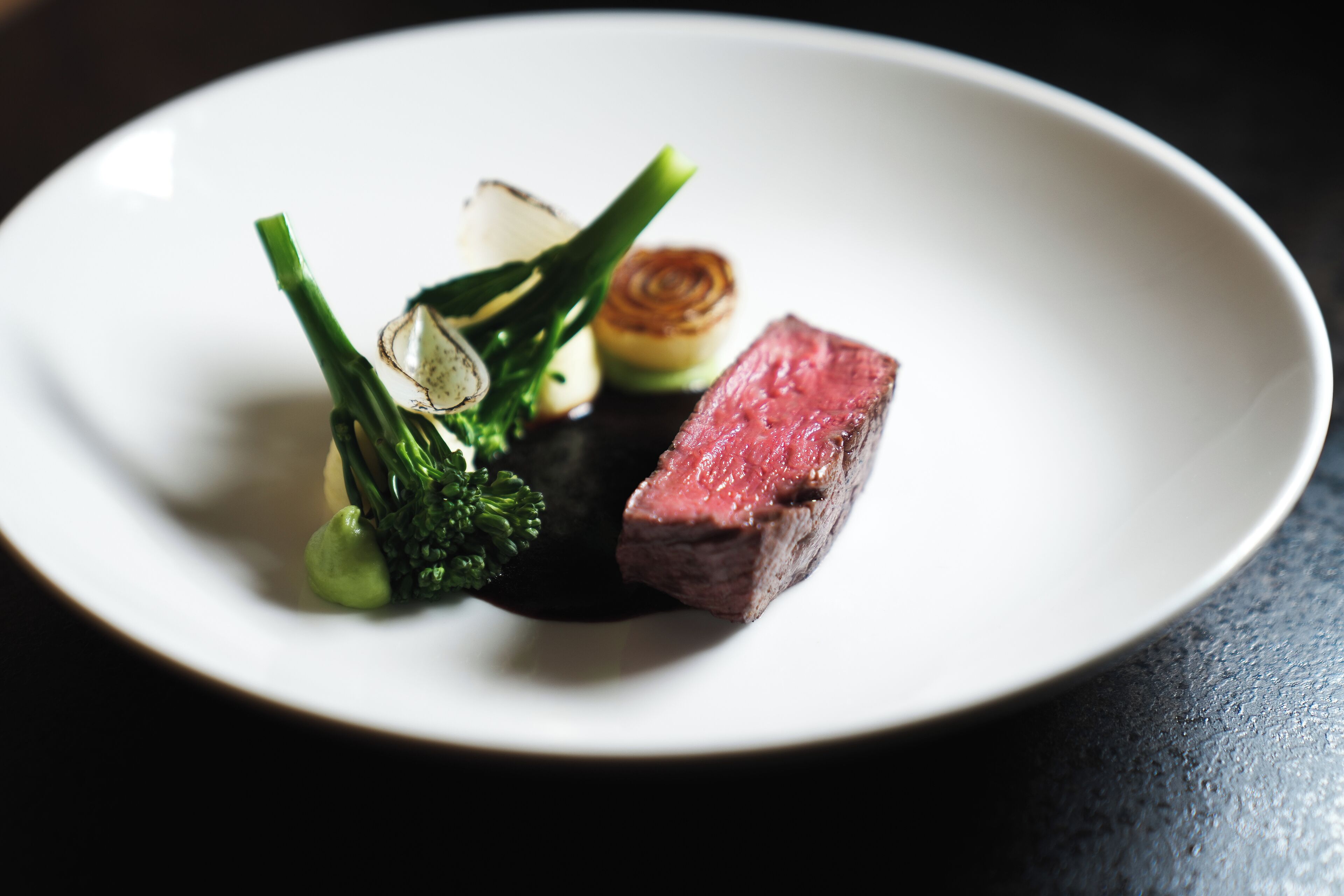 Um prato branco com carne, vegetais verdes e um pedaço pequeno de cebola.