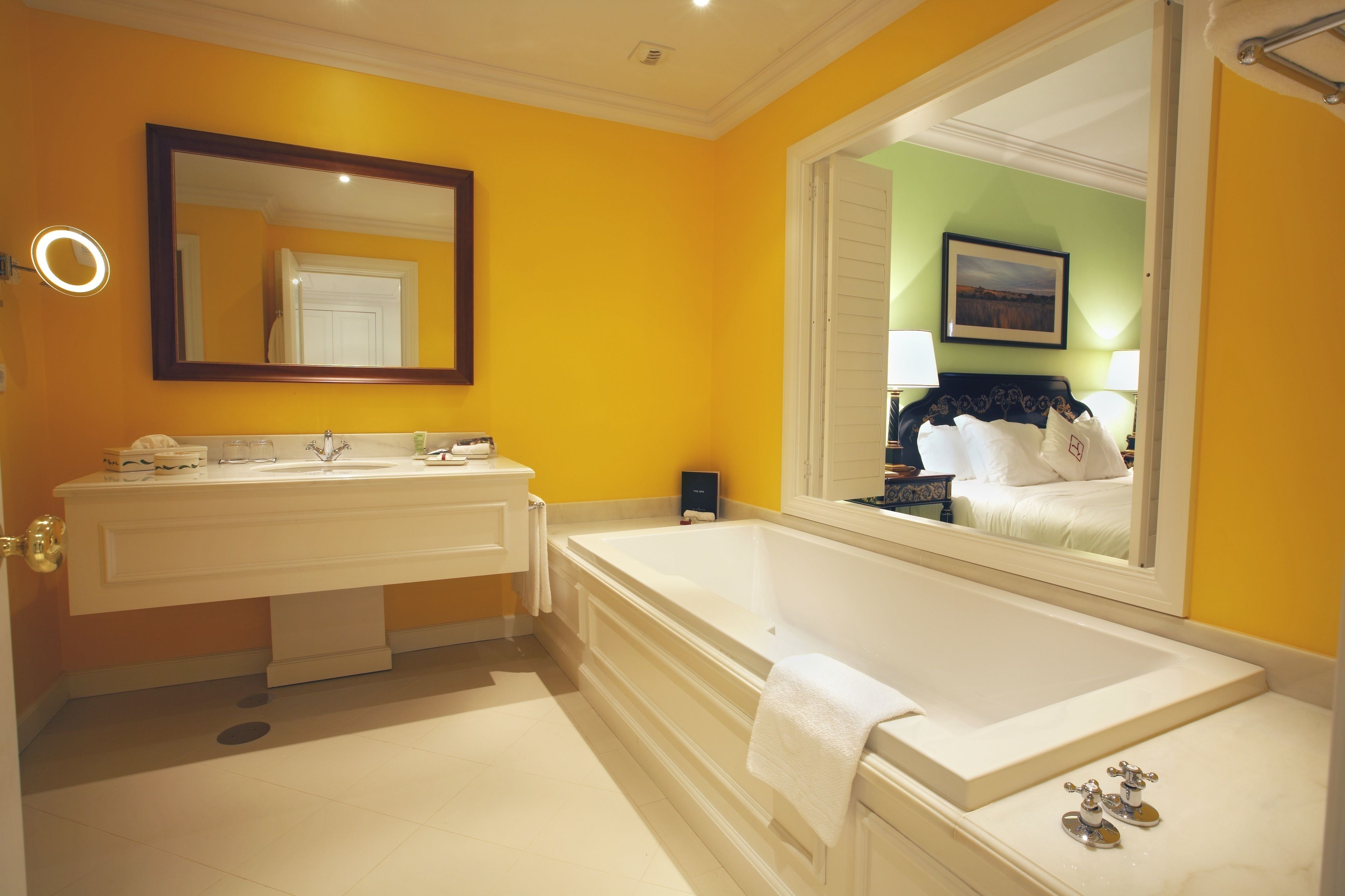 Casa de banho com uma banheira grande, duas pias, um espelho e vista para um quarto com uma cama e um quadro.