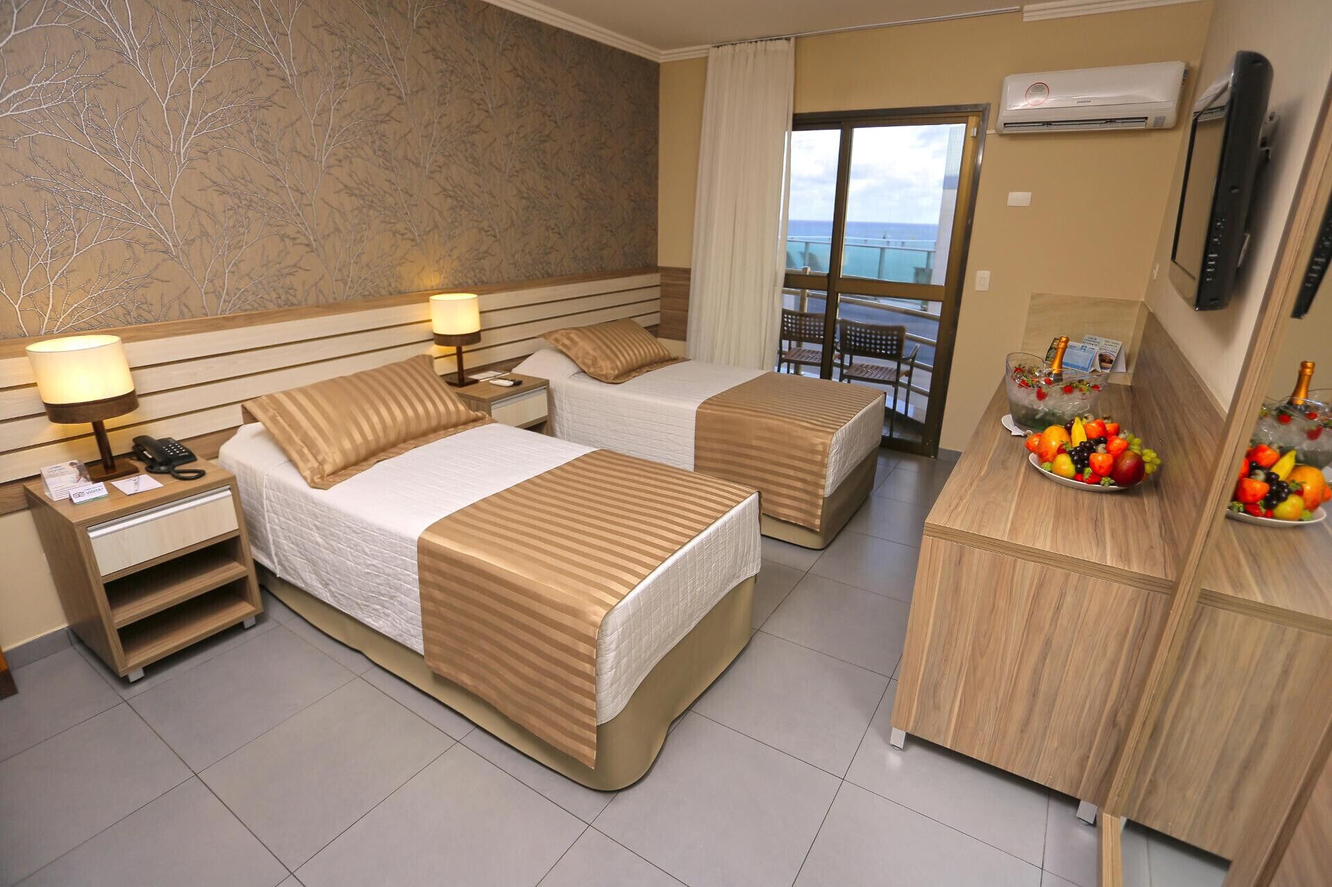 Quarto de hotel com duas camas, uma secretária com uma cesta de frutas e vista para o mar.