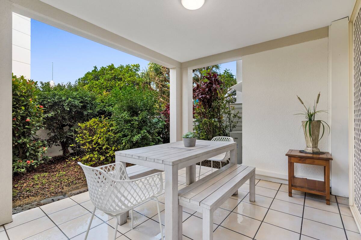 Um pátio com uma mesa e cadeiras brancas, um banco de madeira e uma pequena planta sobre uma mesinha.