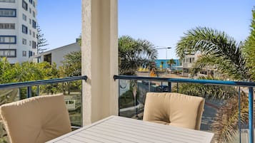 Varanda com duas cadeiras bege acolchoadas e uma mesa branca, com vista para uma rua com palmeiras e edifícios.