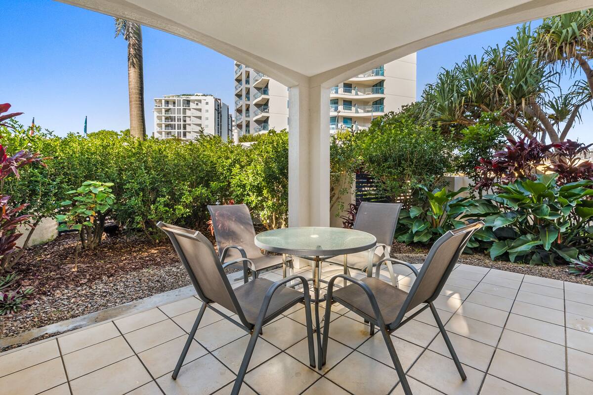 Um pátio externo com uma mesa redonda de vidro e quatro cadeiras, cercado por vegetação exuberante e vista para edifícios modernos ao fundo.