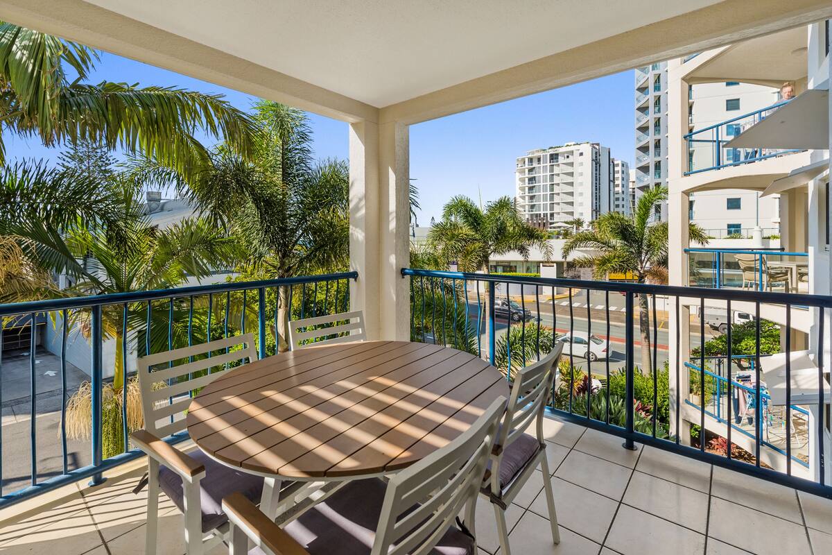 Uma varanda com mesa e cadeiras, com vista para uma rua com palmeiras e edifícios.